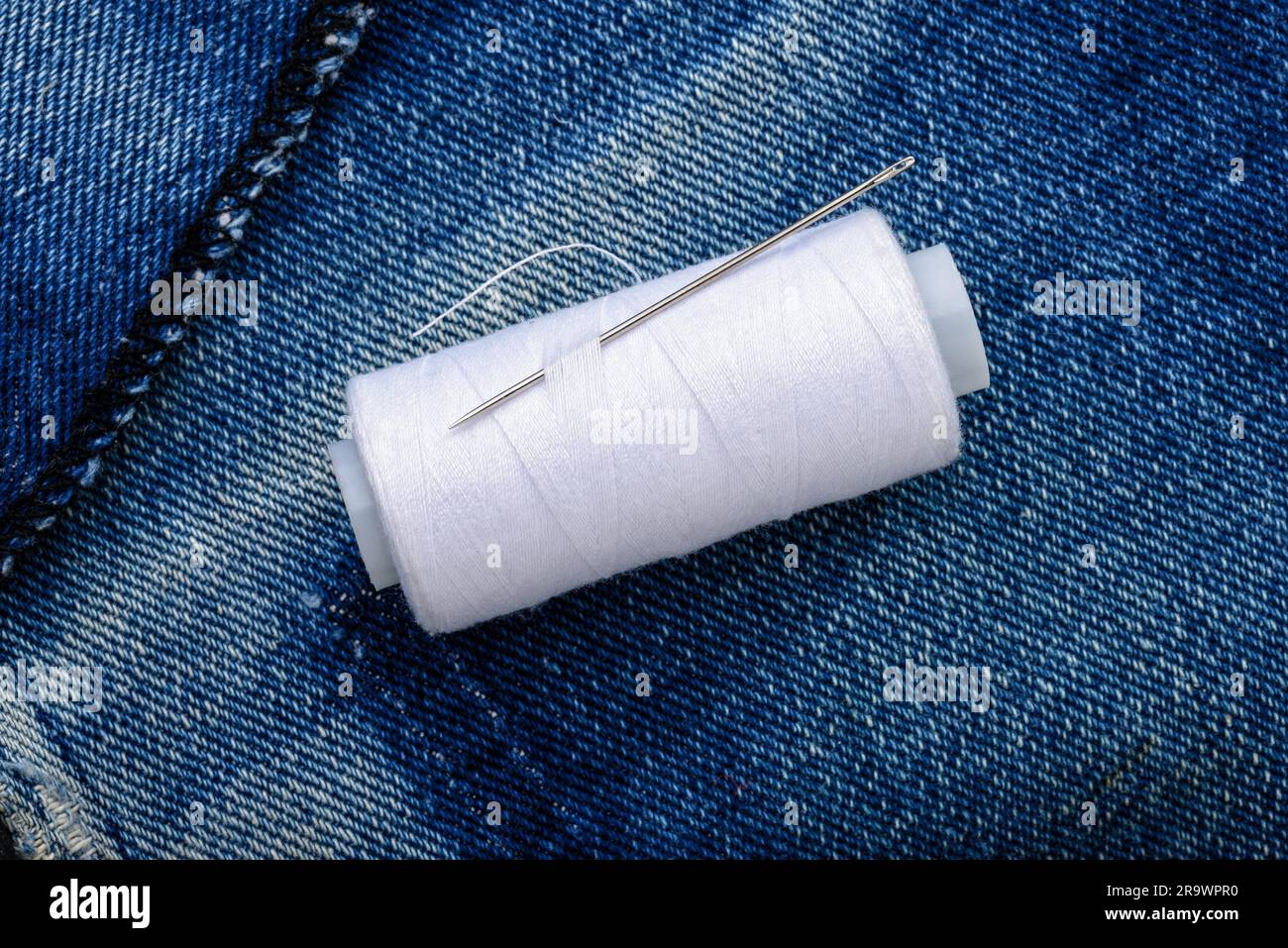 A roll of white thread and a needle on a piece of blue jeans denim ...