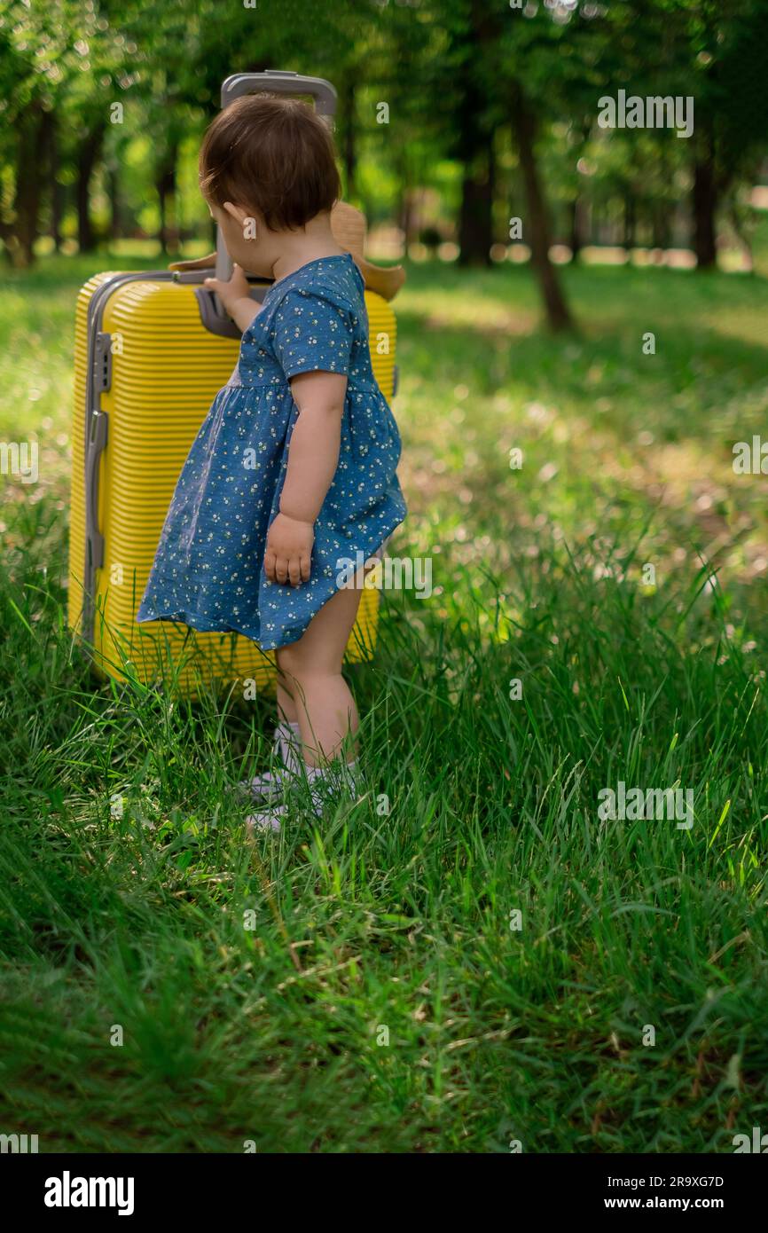 A little happy girl in a blue summer dress stands next to a suitcase ...