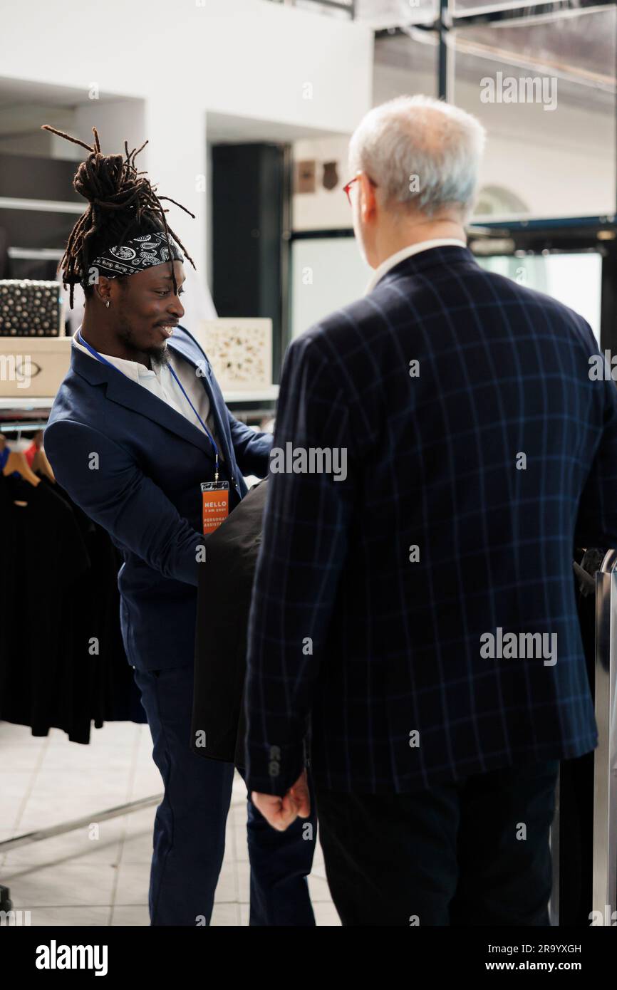 Store employee helping client choosing formal suit in showroom, african ...