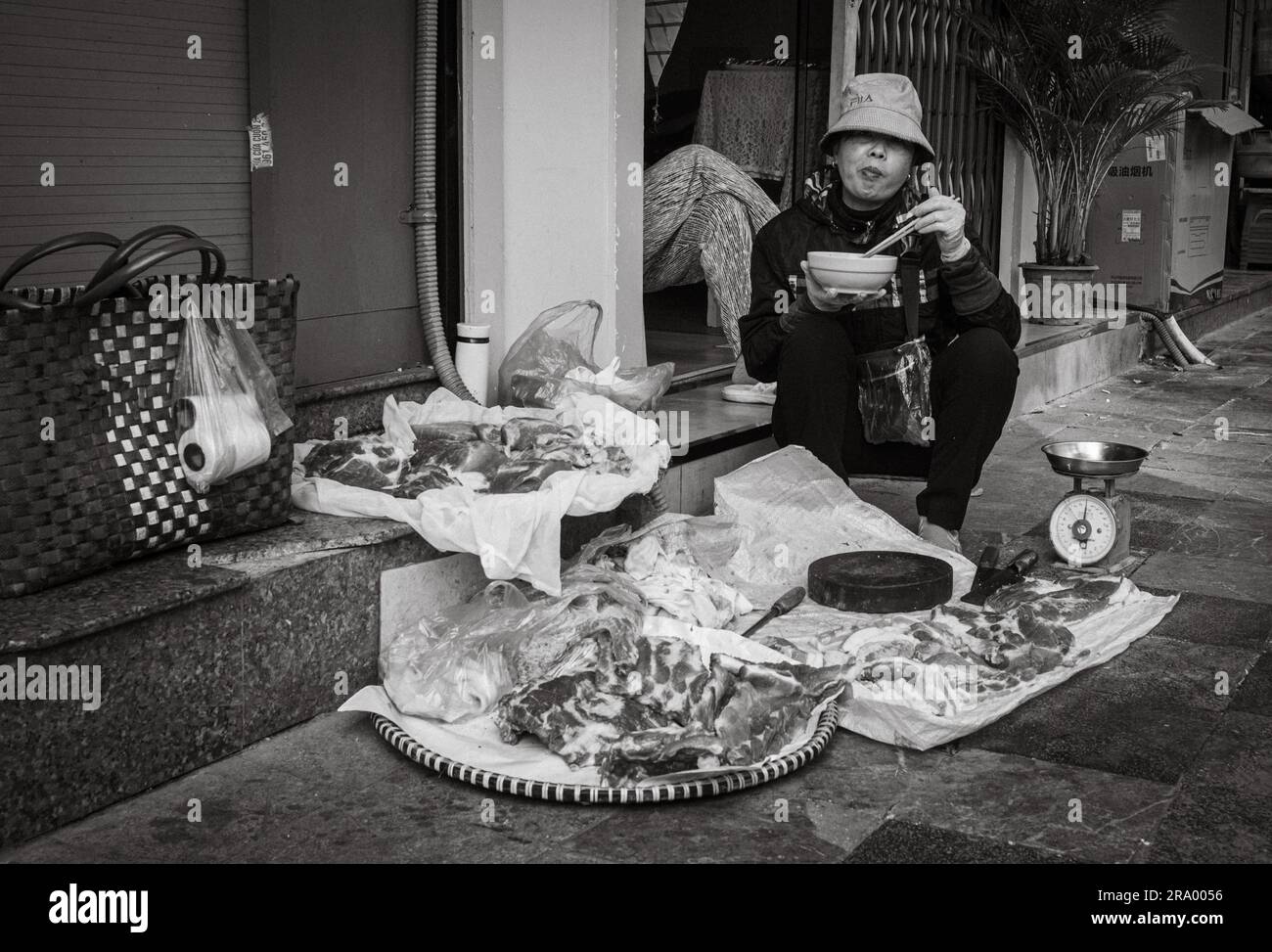A woman eats noodle soup as she sell pork on the pavement in central ...
