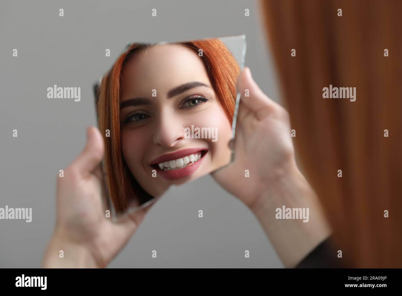 Smiling young woman looking at herself in broken mirror on light grey ...