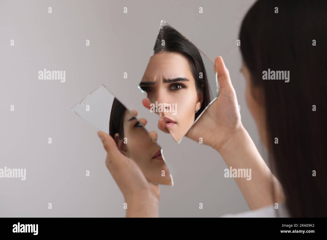 Young woman looking at herself in shard of broken mirror on light grey ...