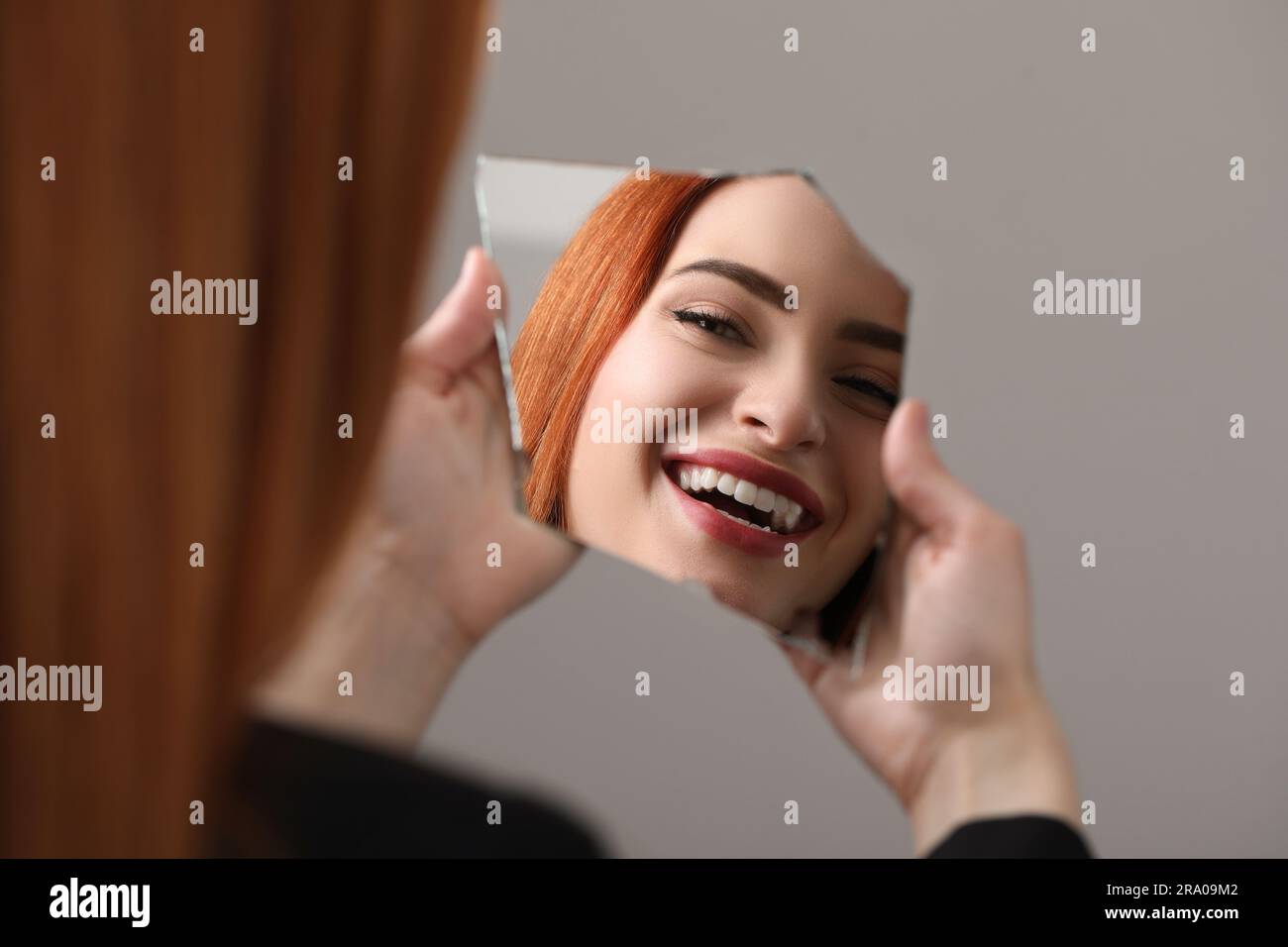 Smiling young woman looking at herself in broken mirror on light grey ...
