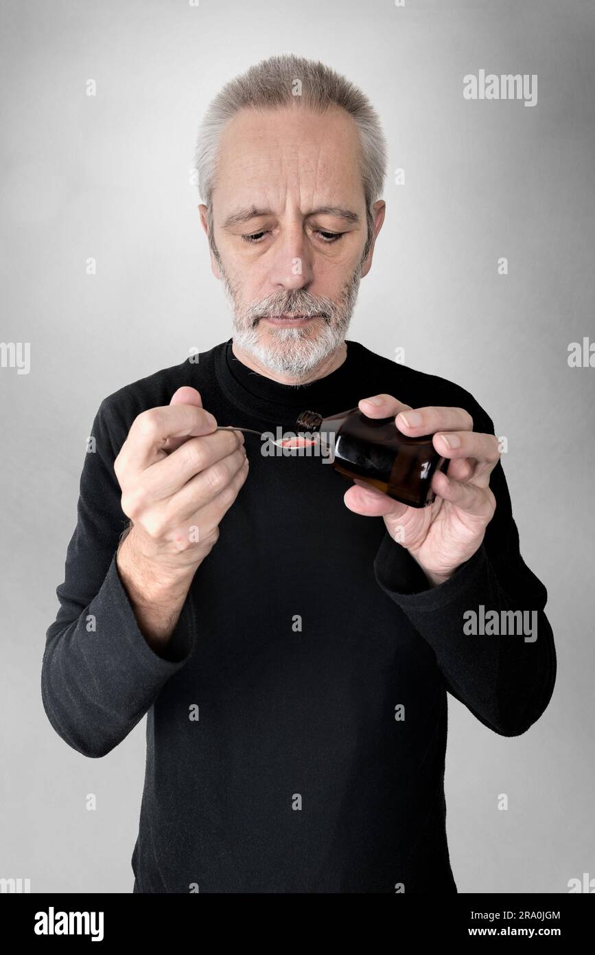 A mature man is pouring cough syrup in a spoon to cure his sore throat ...