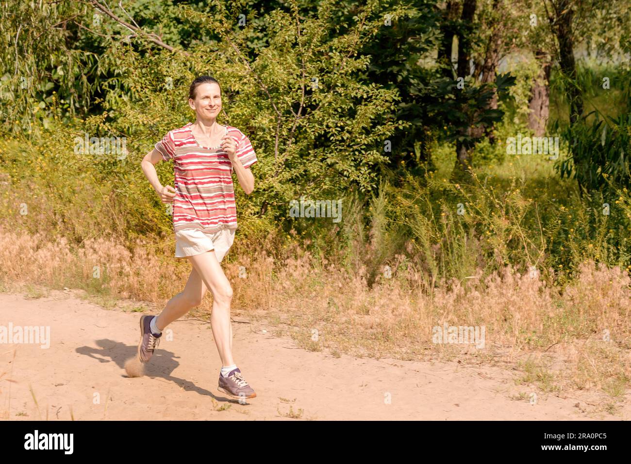 A happy senior woman is running under the sun in the forest during a ...