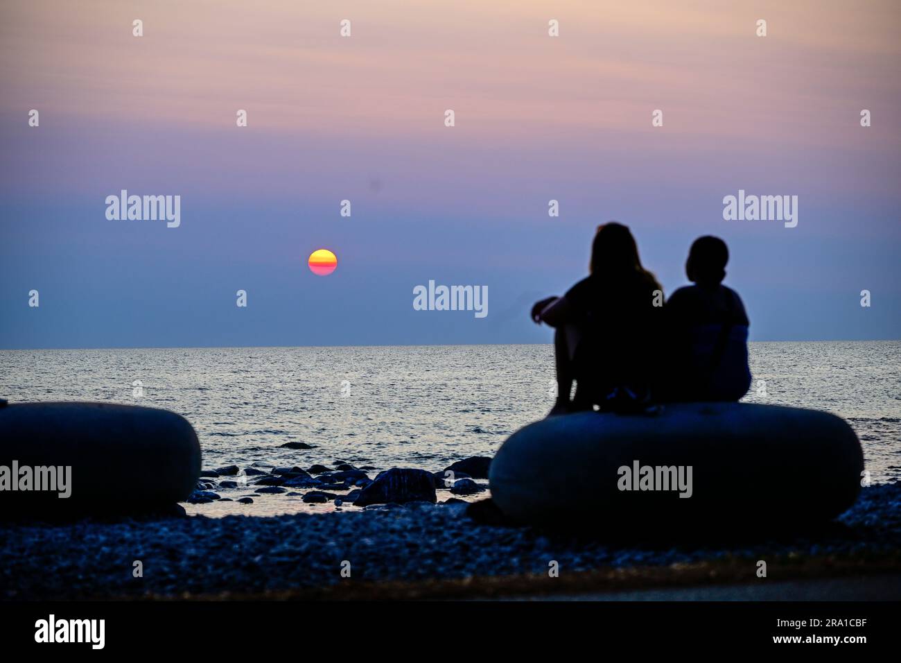 Two people sitting on a large stone on the beach enjoying the sunset in ...