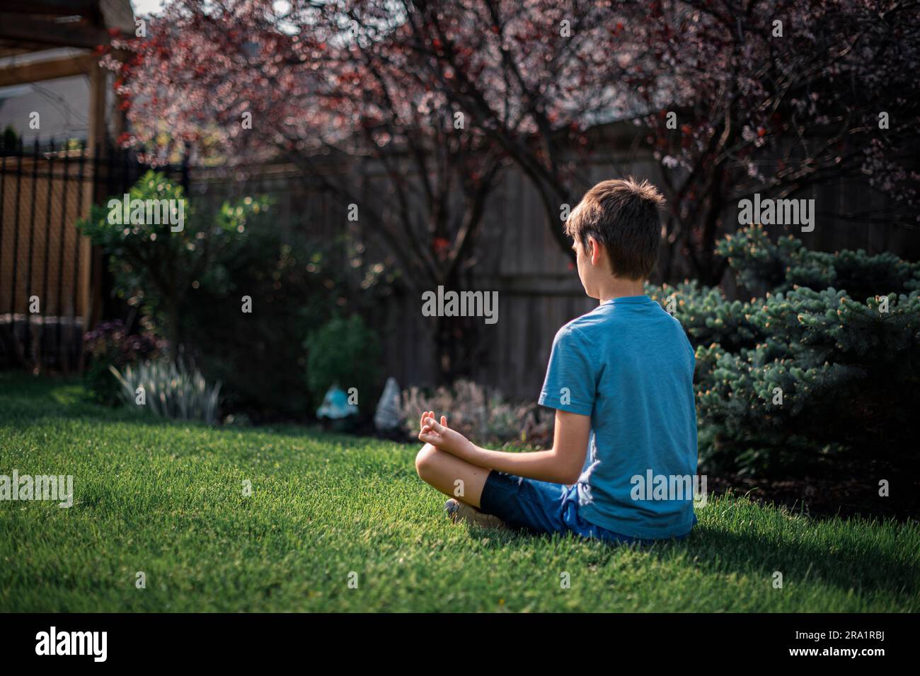 Back view of tween boy sitting on the grass in a garden meditating ...