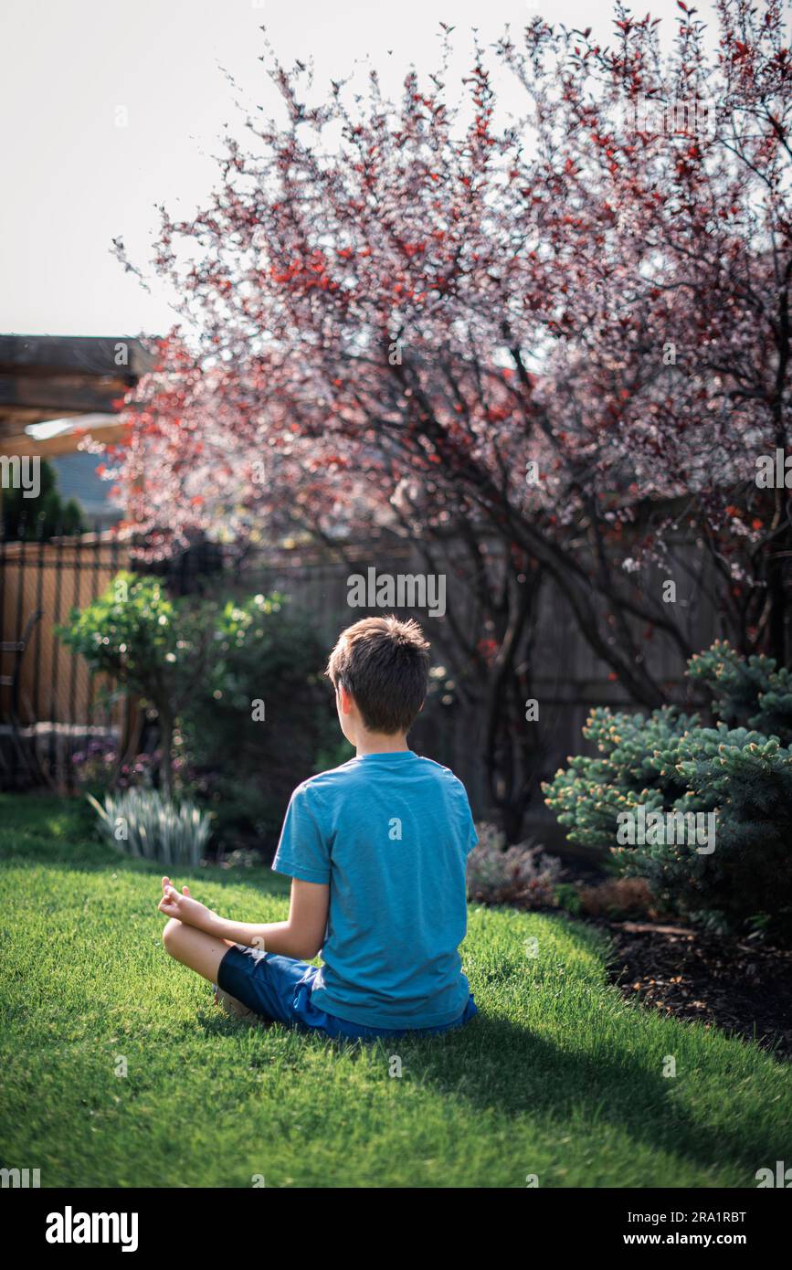 Back view of tween boy sitting on the grass in a garden meditating ...