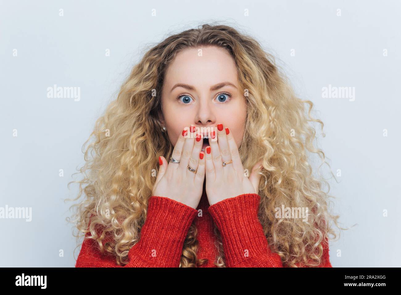 Surprised woman with curly hair, red manicure, covers mouth in shock, isolated on white ...