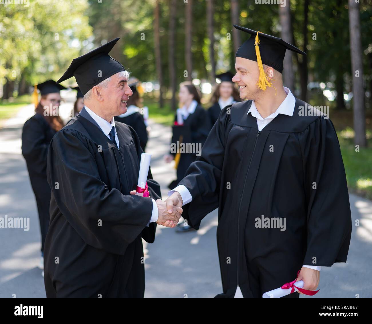 A group of graduates in robes outdoors. An elderly man and a young guy ...