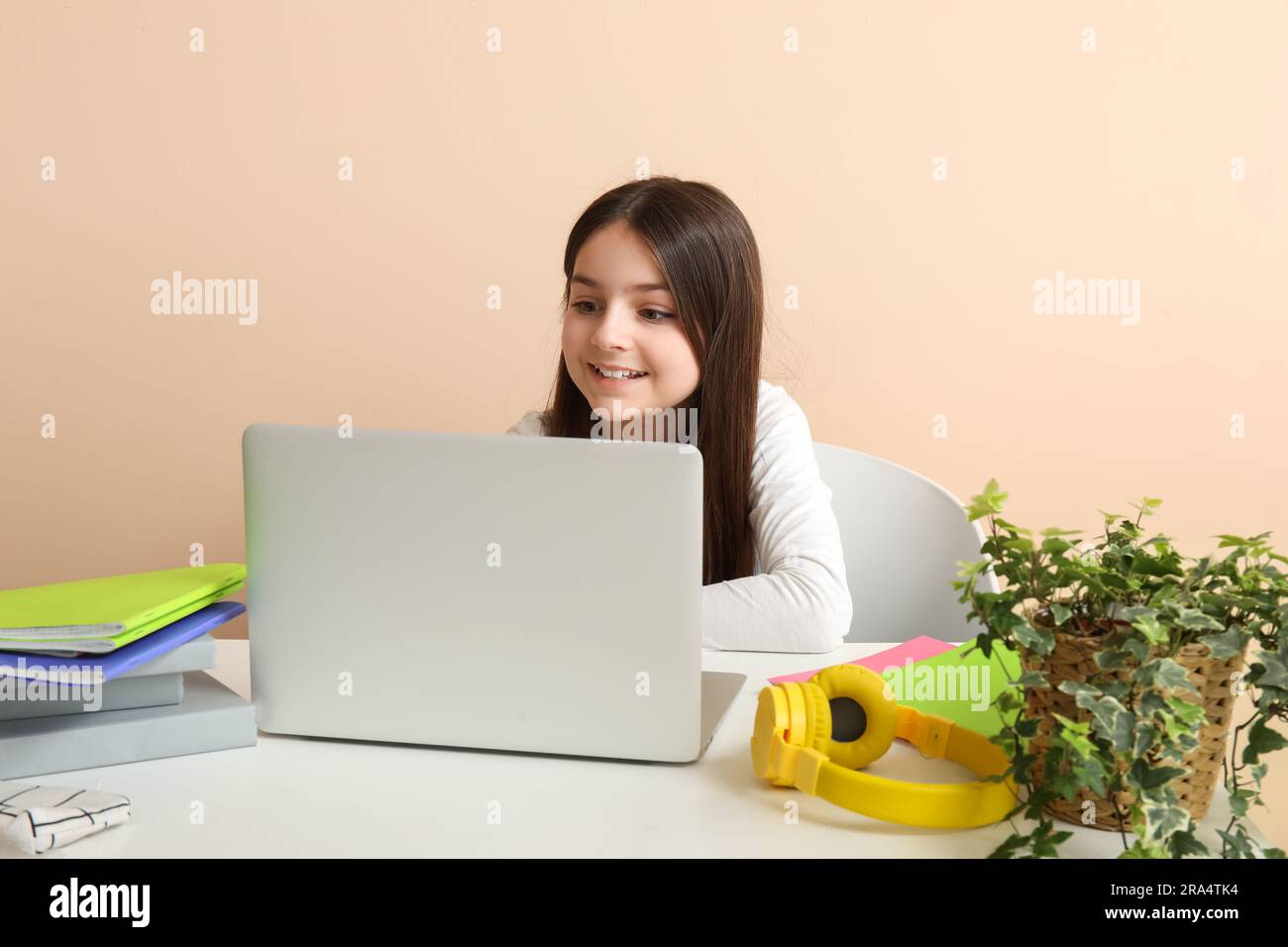 Little girl with laptop doing lessons at table on beige background ...