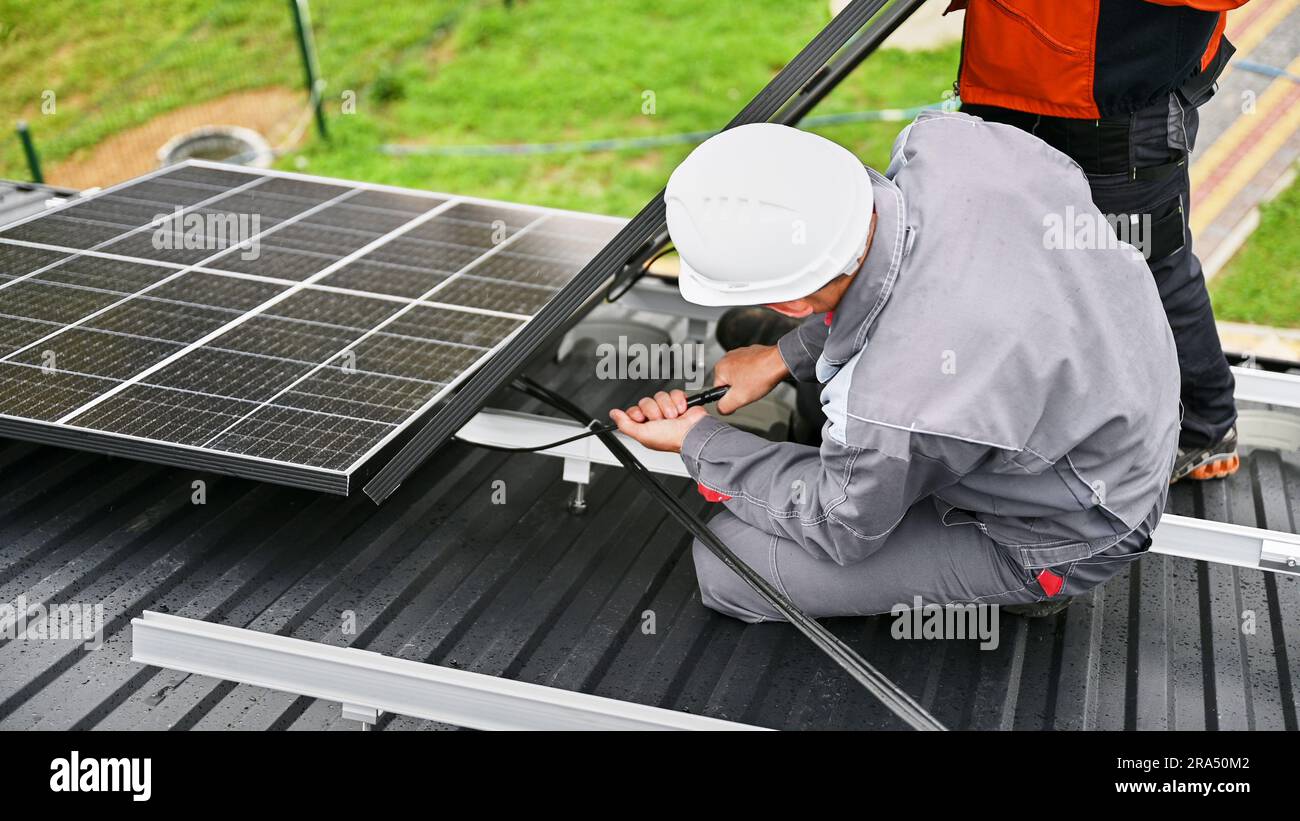 Mounters connecting cables while installing photovoltaic solar panels on roof of house. Workers ...