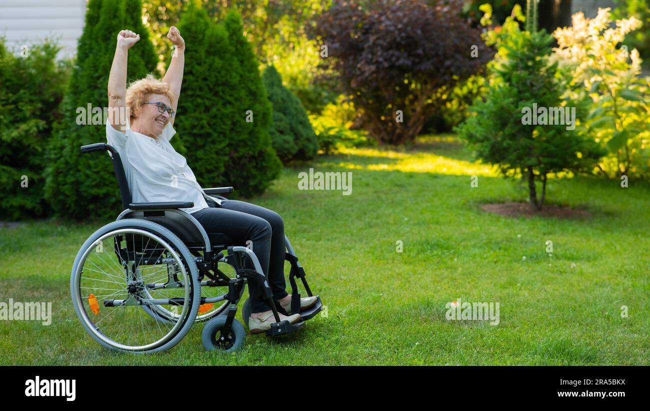 Elderly woman stretches her arms up while sitting in a wheelchair on a ...