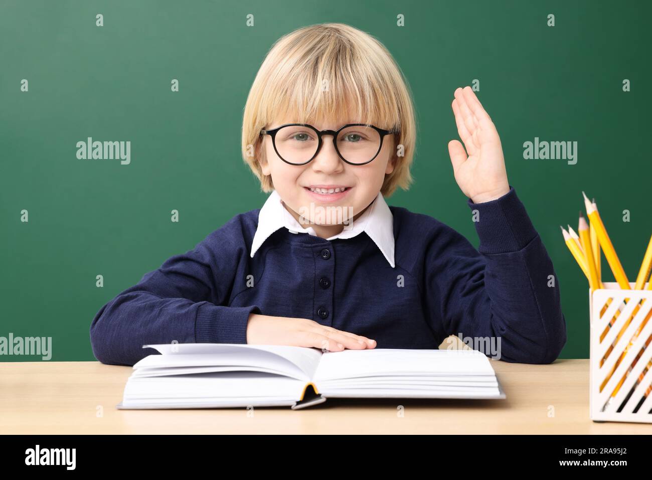 Happy little school child raising hand while sitting at desk with books ...