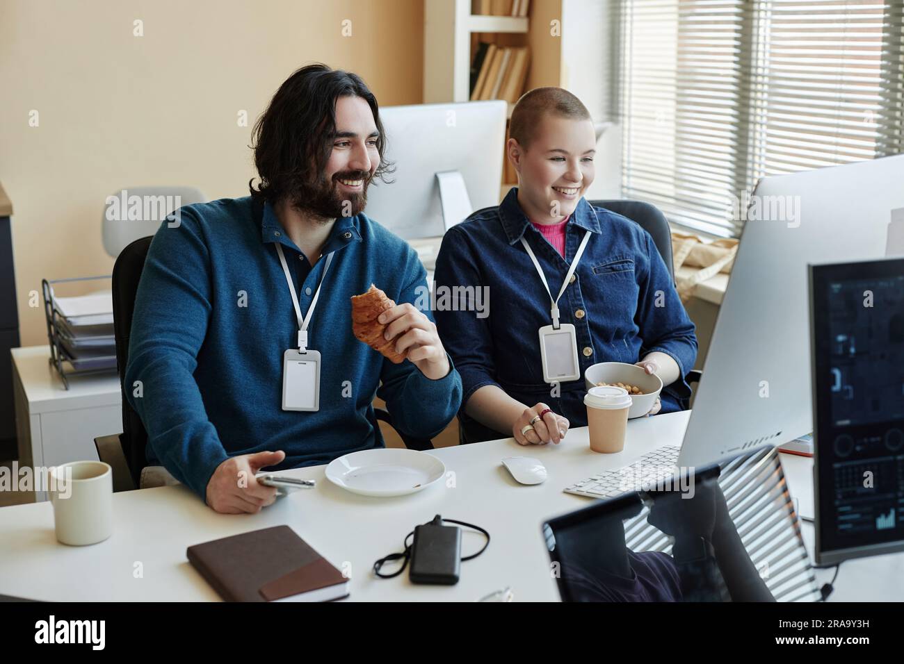 Two happy young office managers having snacks at lunch break while ...