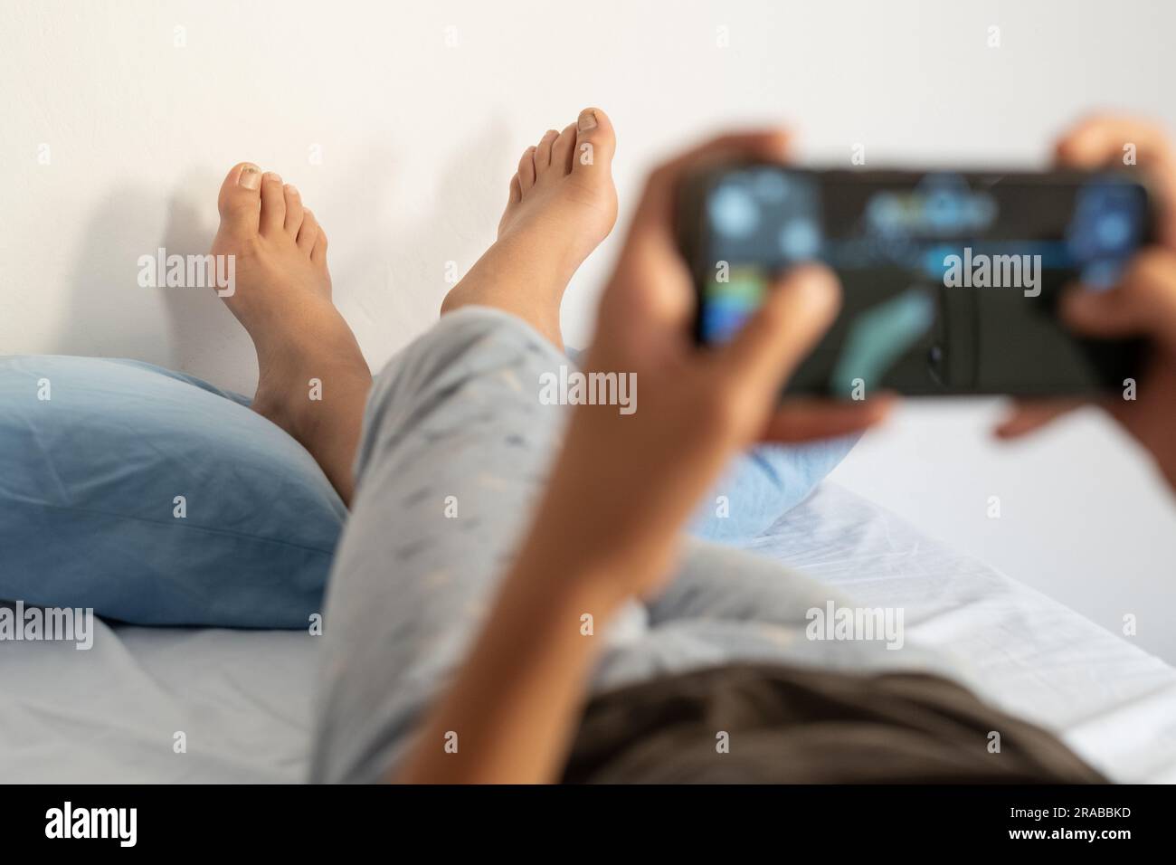 Barefoot teenage boy with crossed legs resting on pillow using phone ...