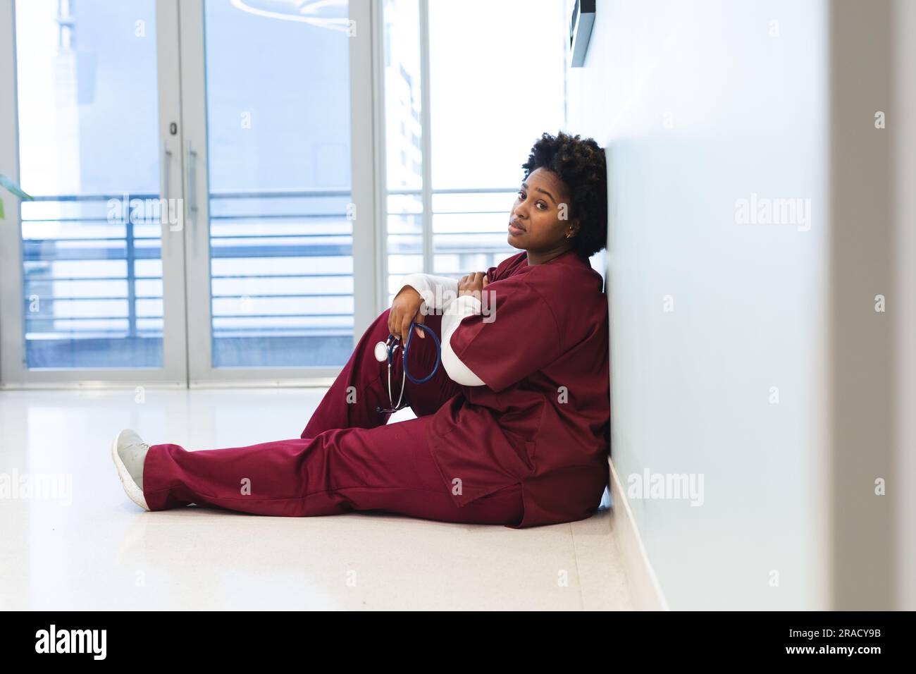 Tired african american female doctor wearing scrubs, sitting on floor ...