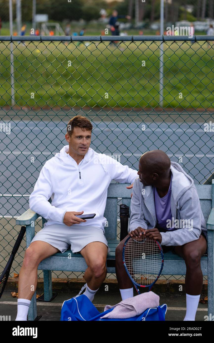 Happy diverse male tennis players sitting on bench talking at outdoor ...