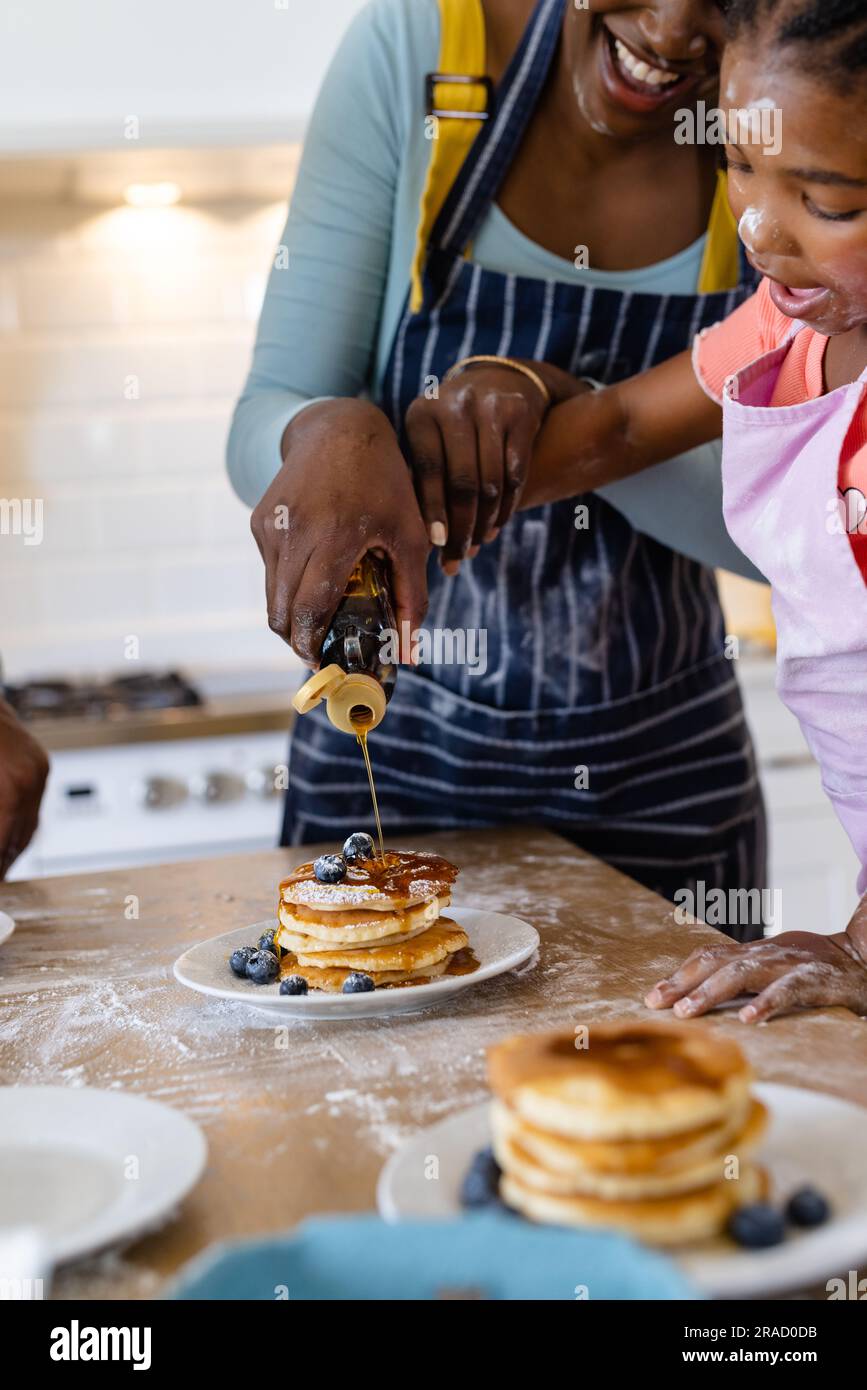 Happy african american mother and daughter pouring honey on pancakes in plate at home Stock ...