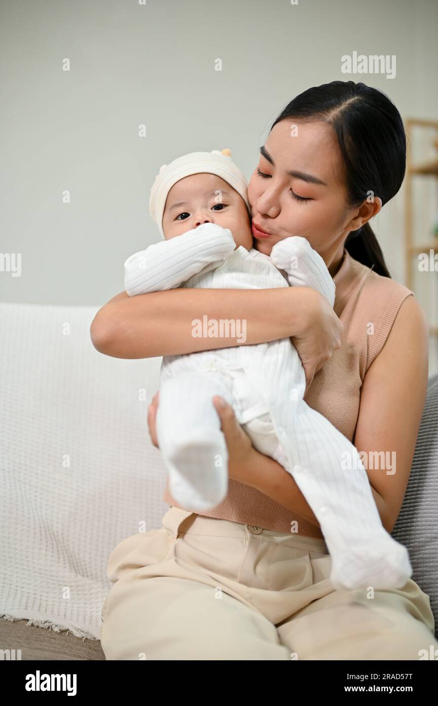 Beautiful Asian Mother holding her baby boy while relaxing on the sofa, spending time with her ...