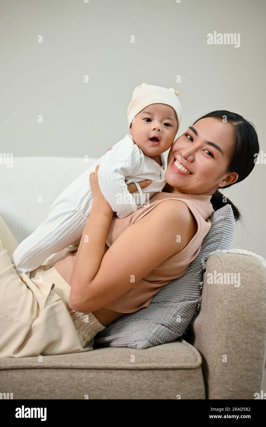 Beautiful Asian Mother holding her baby boy while relaxing on the sofa, spending time with her ...