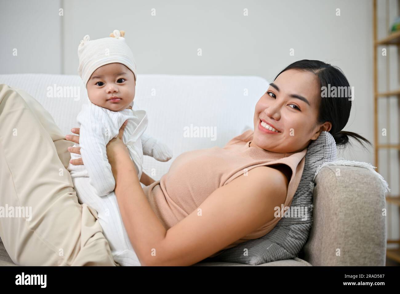 Beautiful Asian Mother holding her baby boy while relaxing on the sofa, spending time with her ...