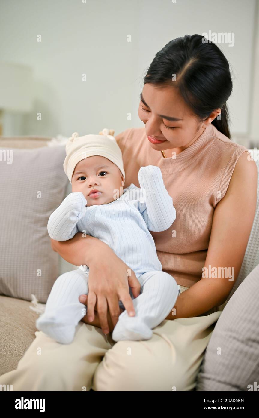 Beautiful Asian Mother holding her baby boy while relaxing on the sofa, spending time with her ...