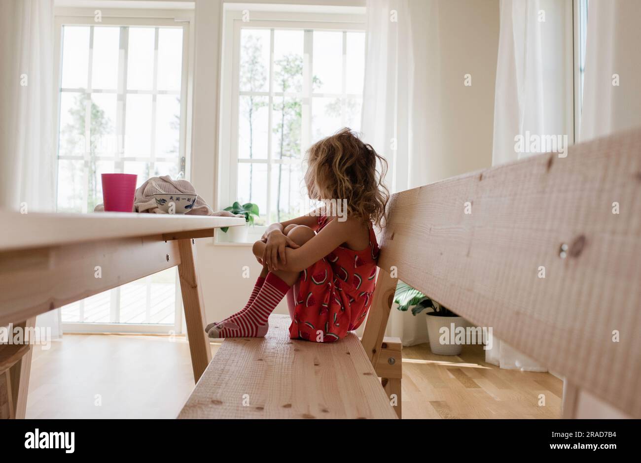young girl sat on a bench at home hugging her legs thinking on her own ...