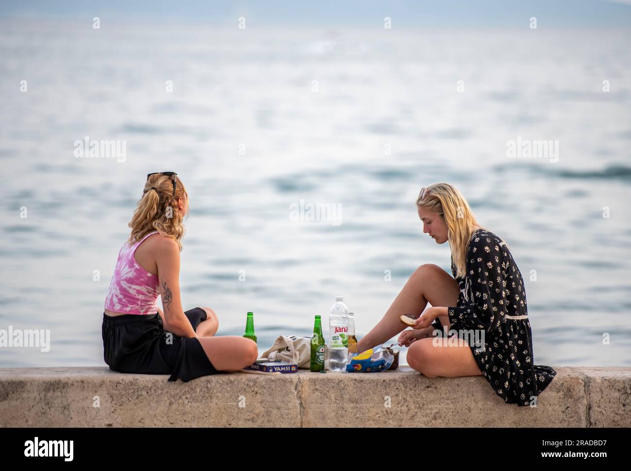 two attractive young women sitting on wall at the seaside sharing ...