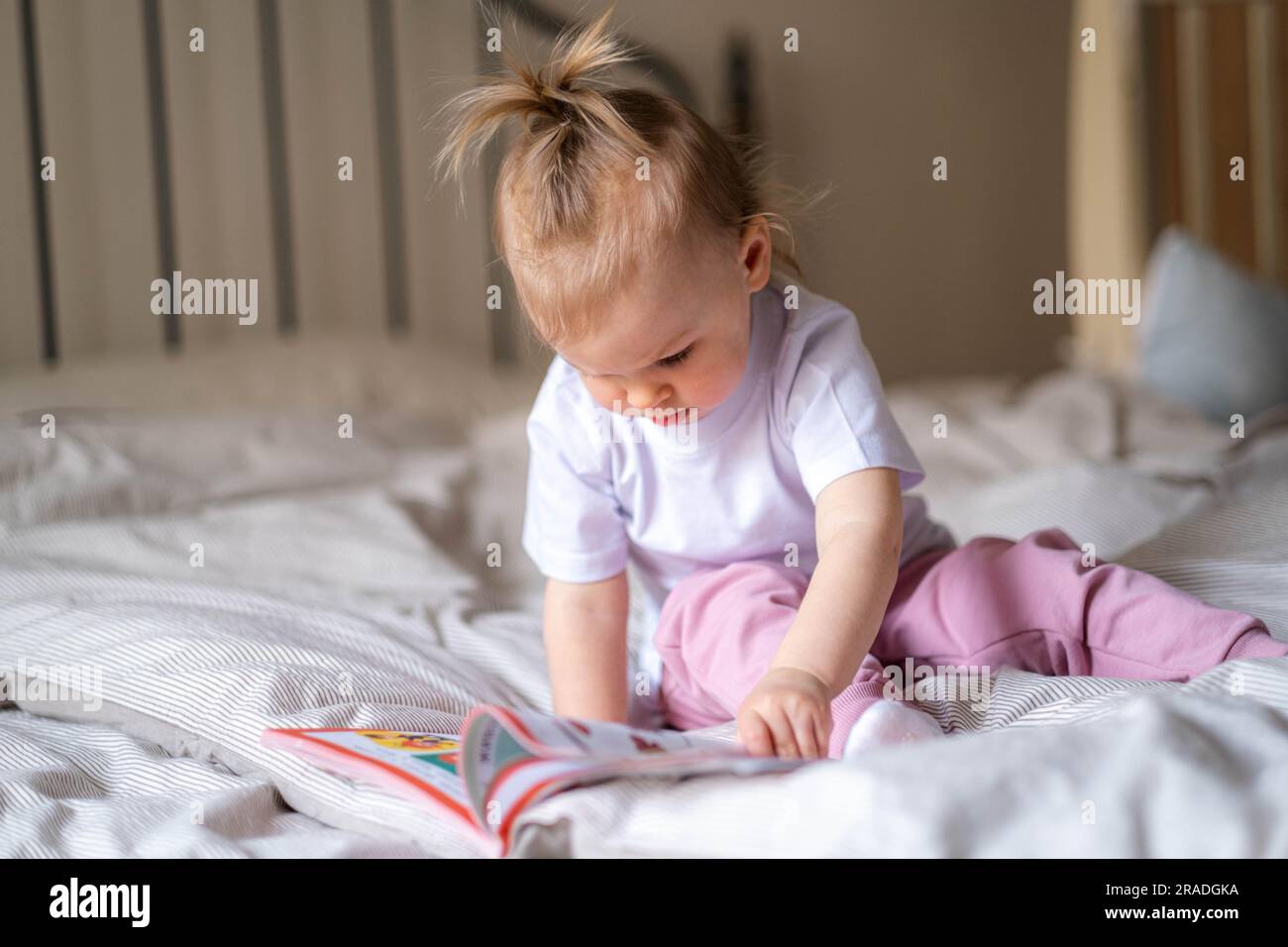 little baby girl reading, looking at pictures in book on bed at home ...