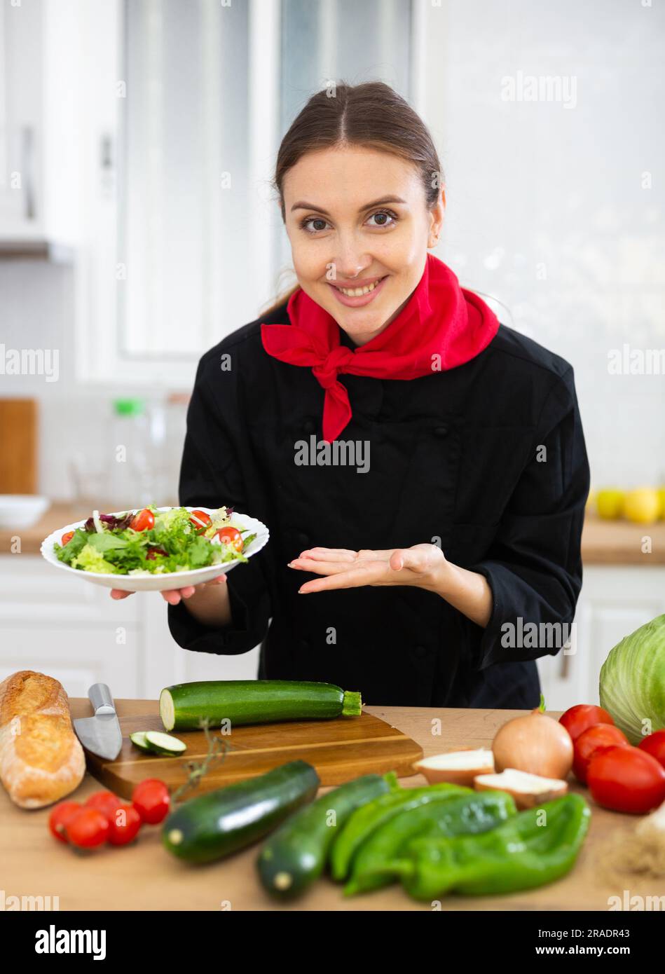 Smiling female chef in black uniform offering plate of vegetable salad ...