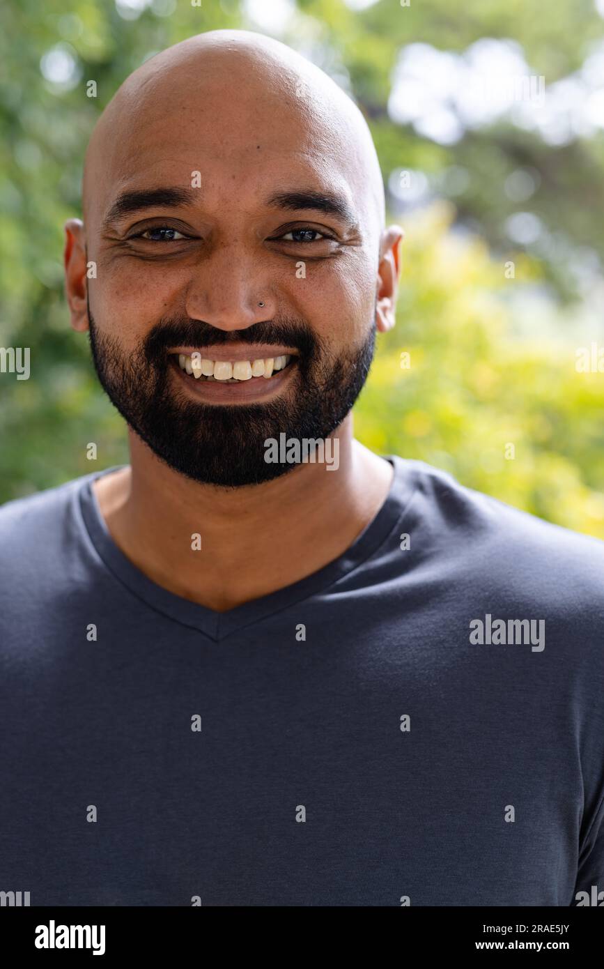 Portrait of smiling bald biracial man with beard standing on sunny balcony in front of treetops ...