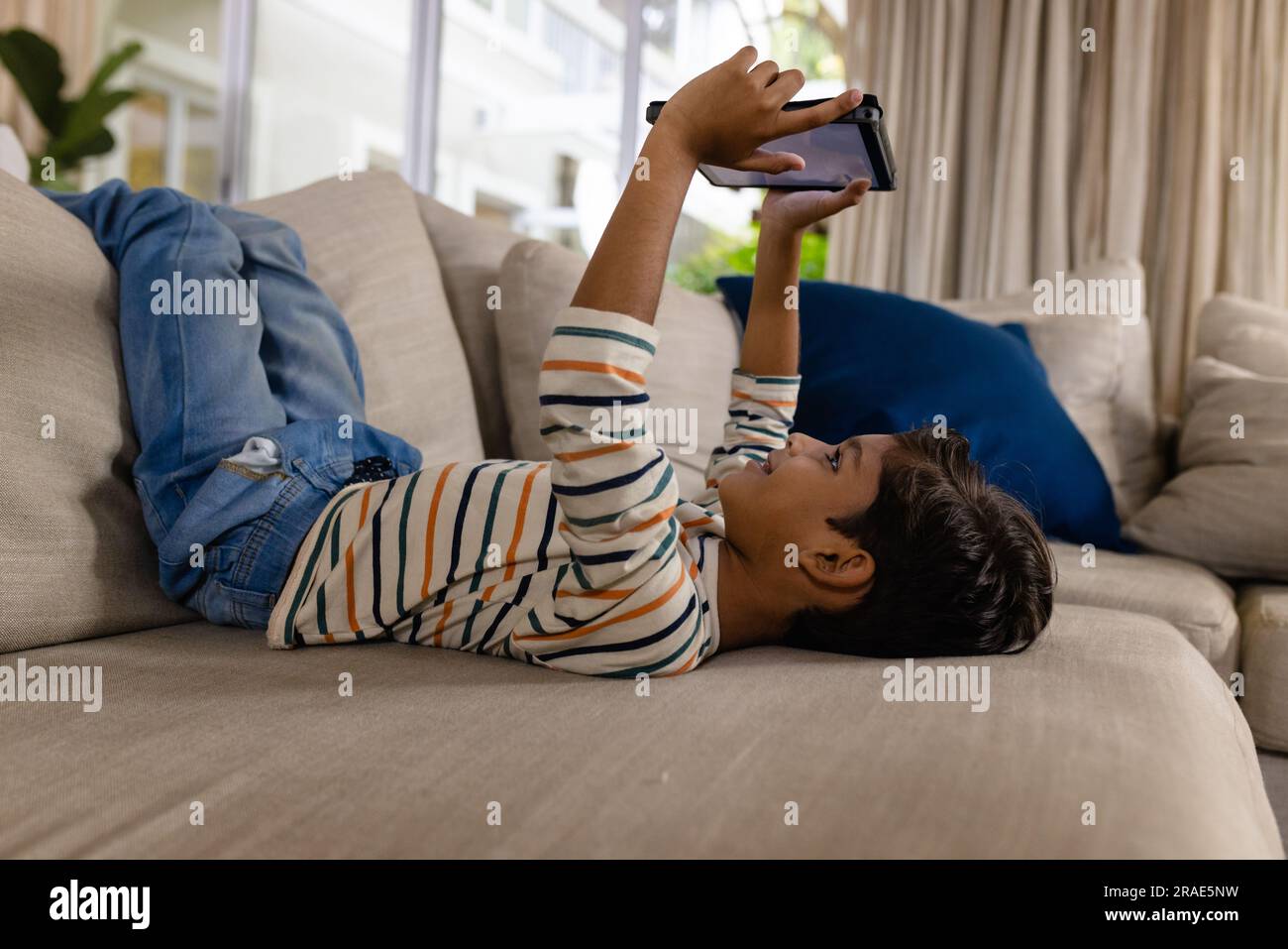Happy biracial boy lying on couch with feet up using tablet in living ...