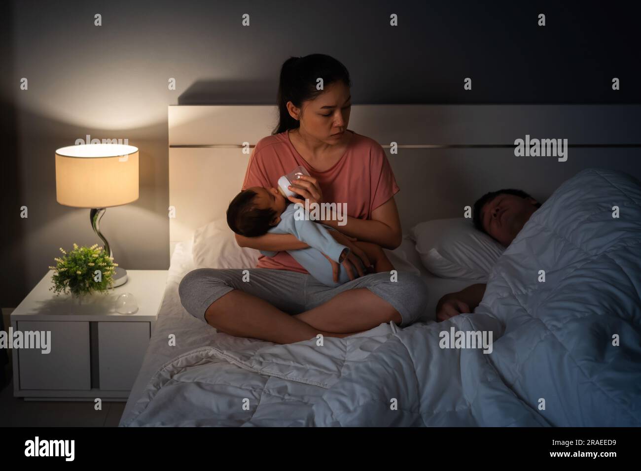 tired mother feeding milk bottle to newborn baby while looking her husband sleeping on a bed at ...