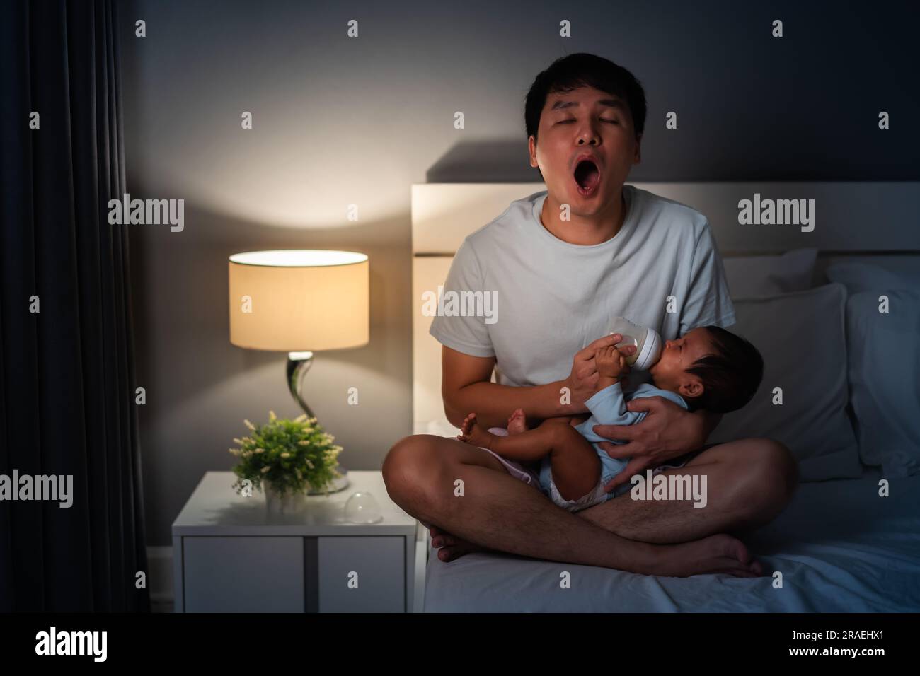 sleepy father yawning and feeding milk bottle to newborn baby on a bed ...