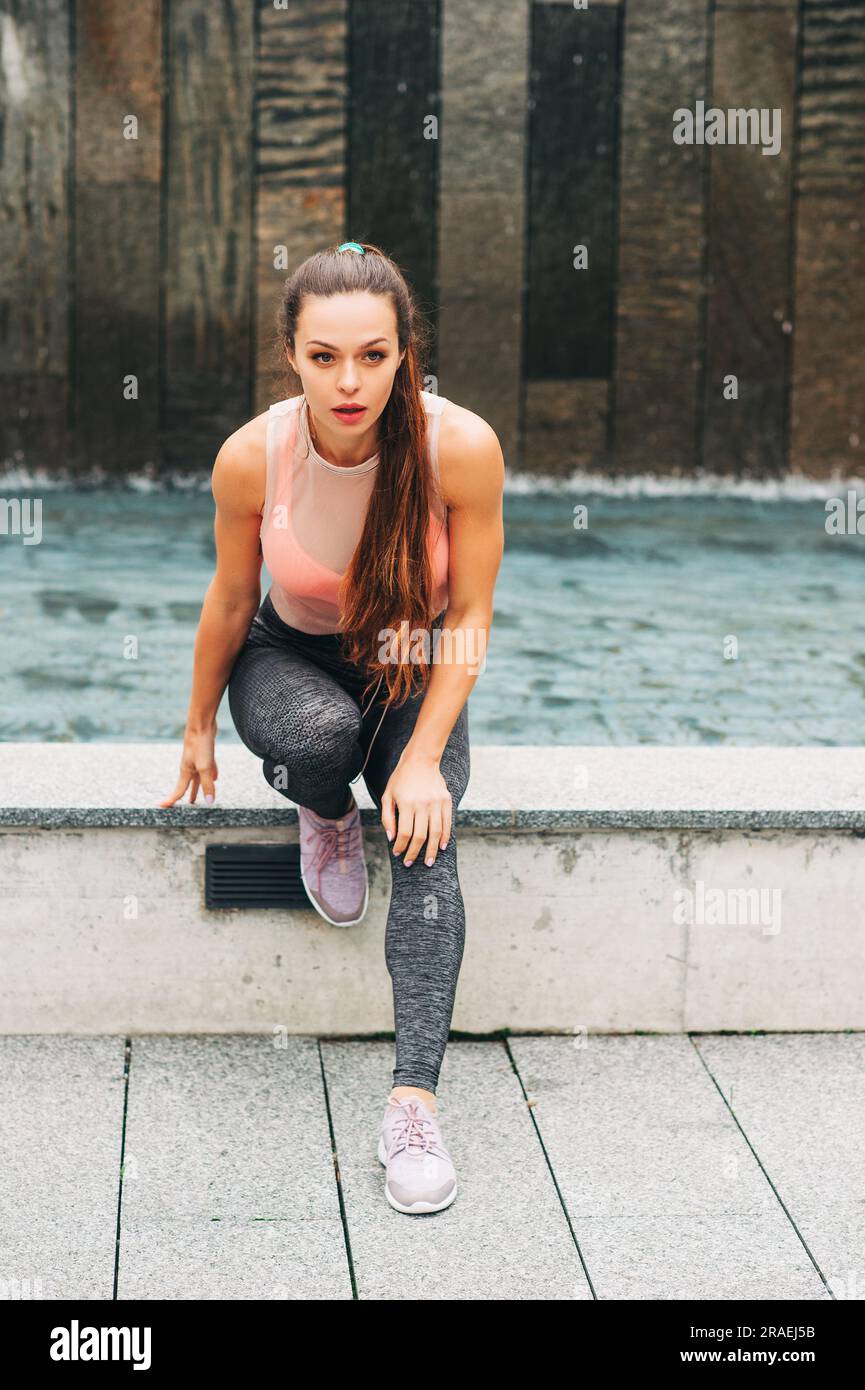 Outdoor portrait of young beautiful fit woman stretching legs, wearing ...