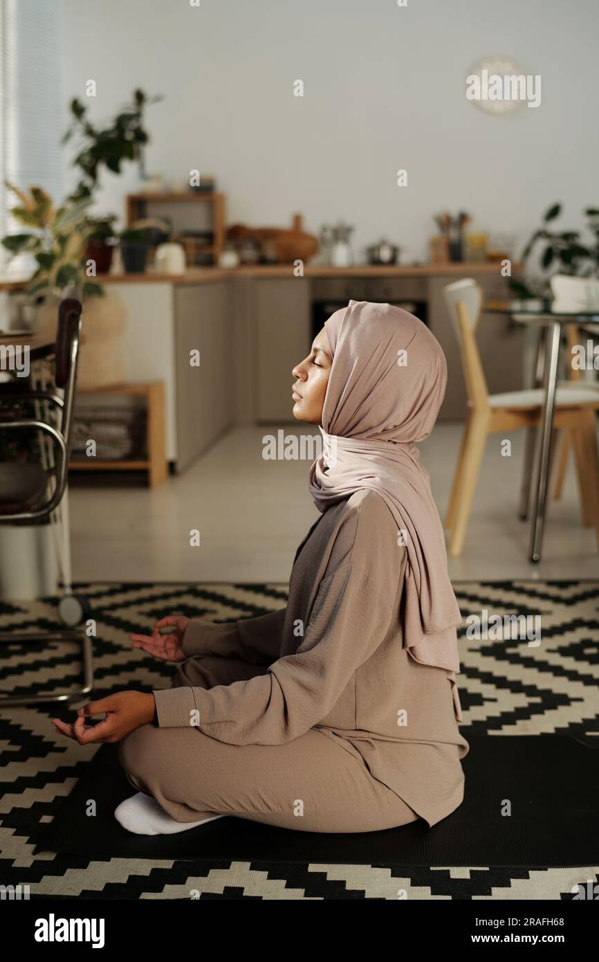 Side view of young Muslim woman sitting in pose of lotus on mat in ...