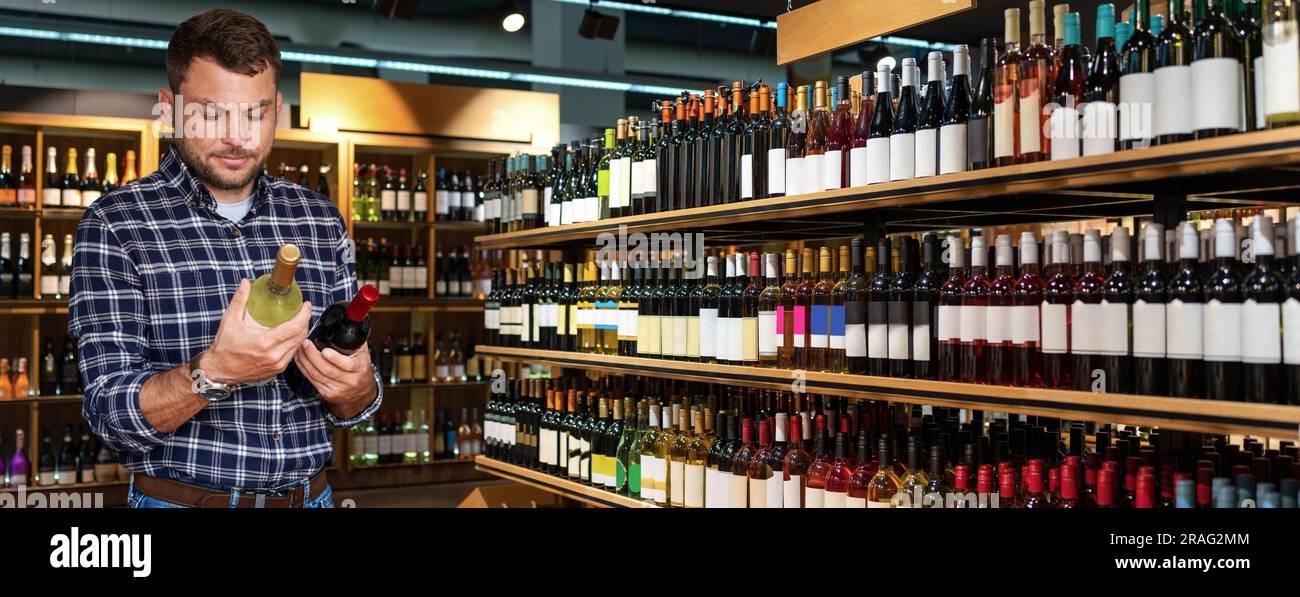 Man customer choosing wine in a liquor store, holding two bottles of ...