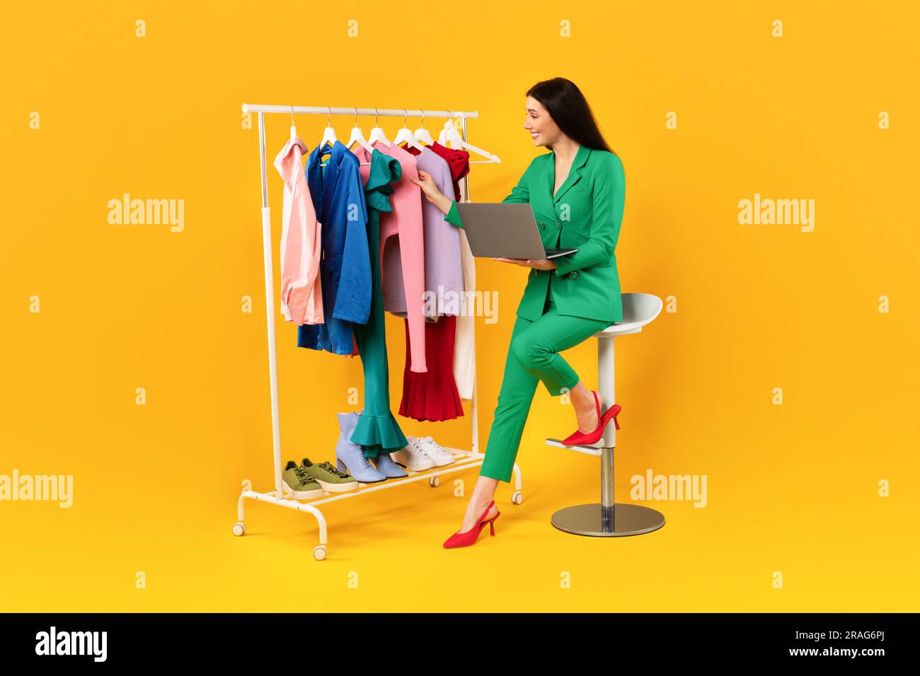 Lovely young european woman with laptop touching clothes on rack ...