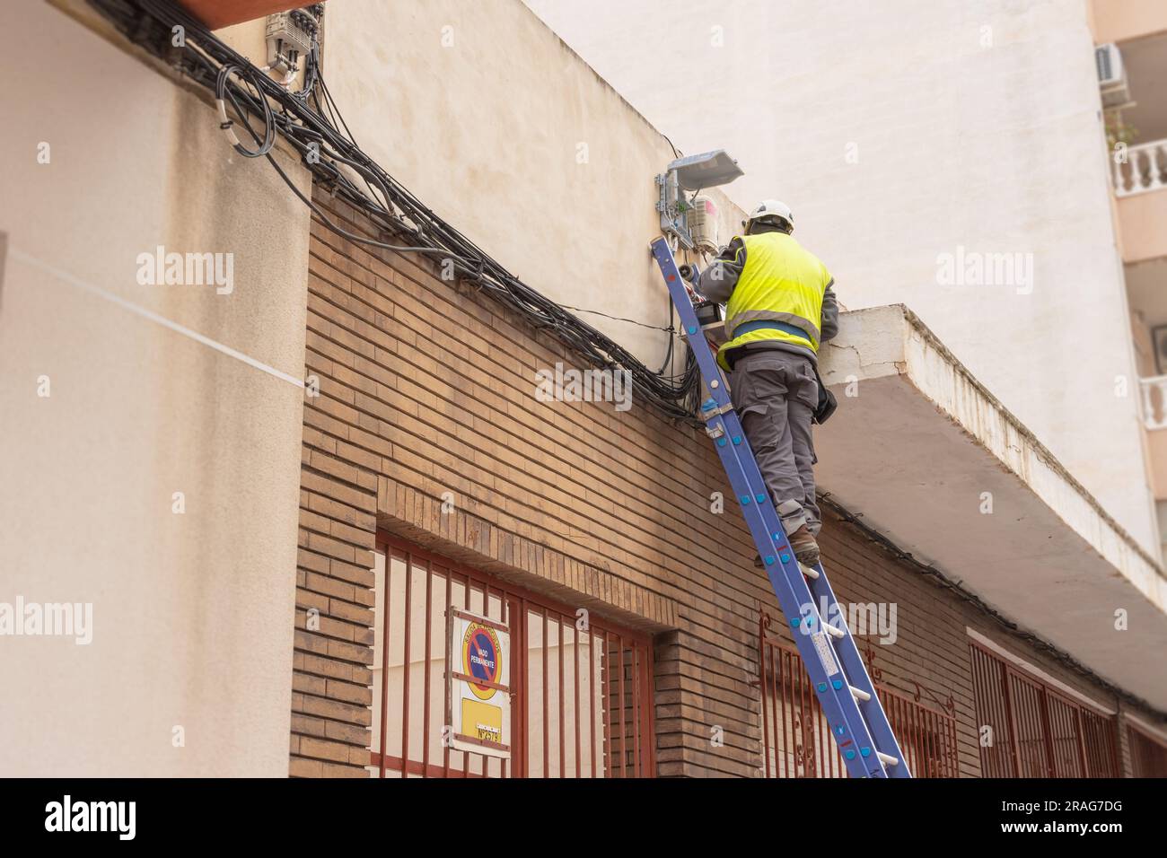 A man in a reflective vest stands on a stepladder doing electrics. A man in work clothes repairs ...