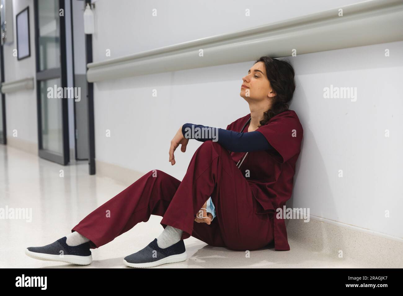 Tired caucasian female doctor wearing scrubs, sitting on floor in ...