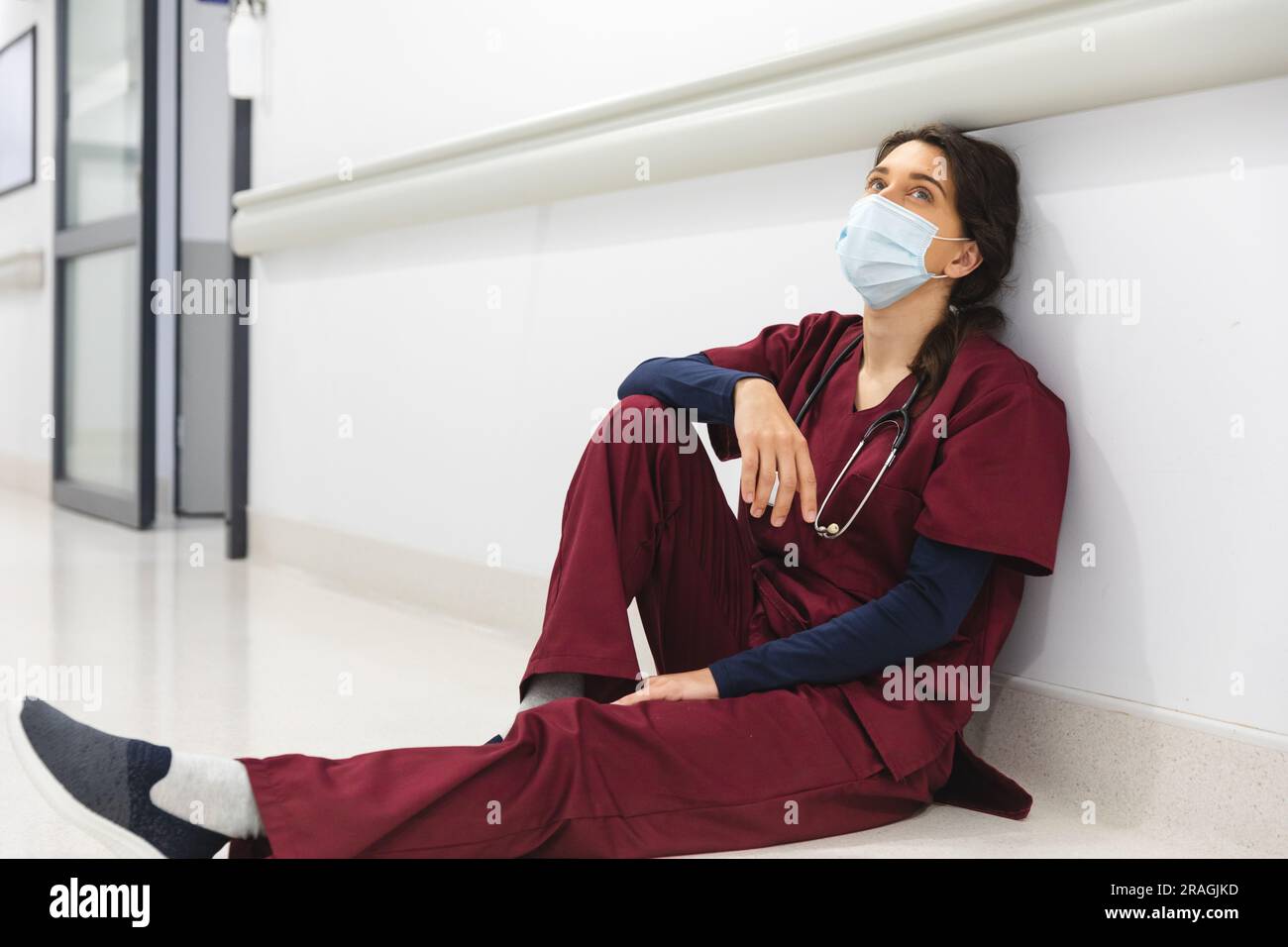Tired caucasian female doctor wearing scrubs and face mask, sitting on ...