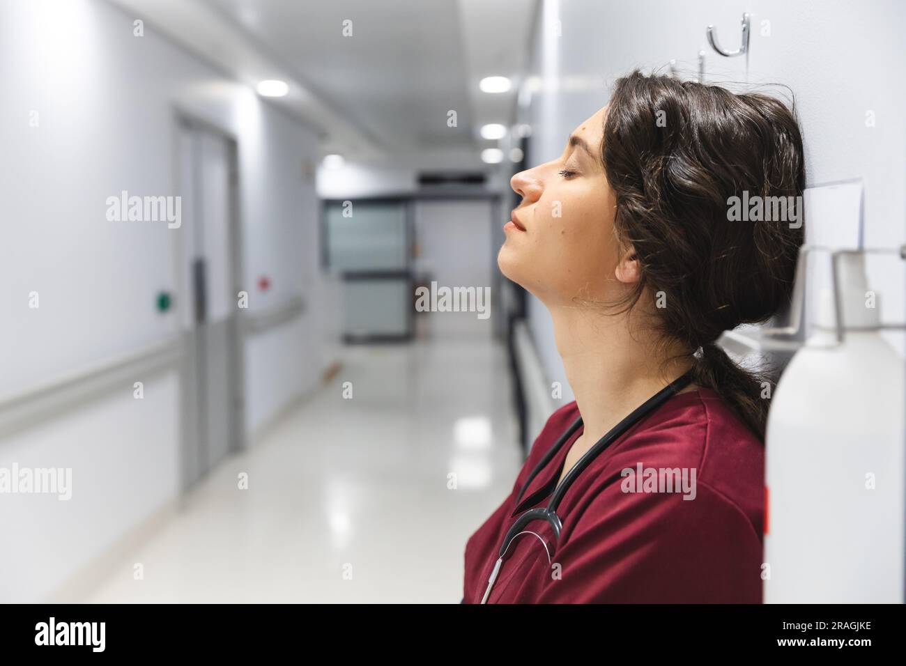 Tired caucasian female doctor wearing scrubs, leaning on wall in ...