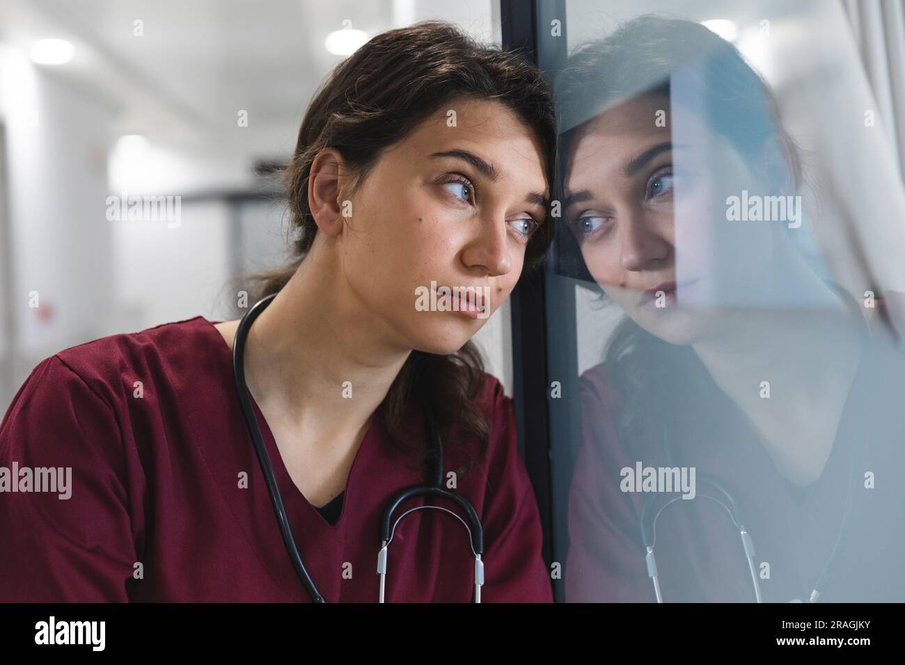 Tired caucasian female doctor wearing scrubs, leaning on wall in ...