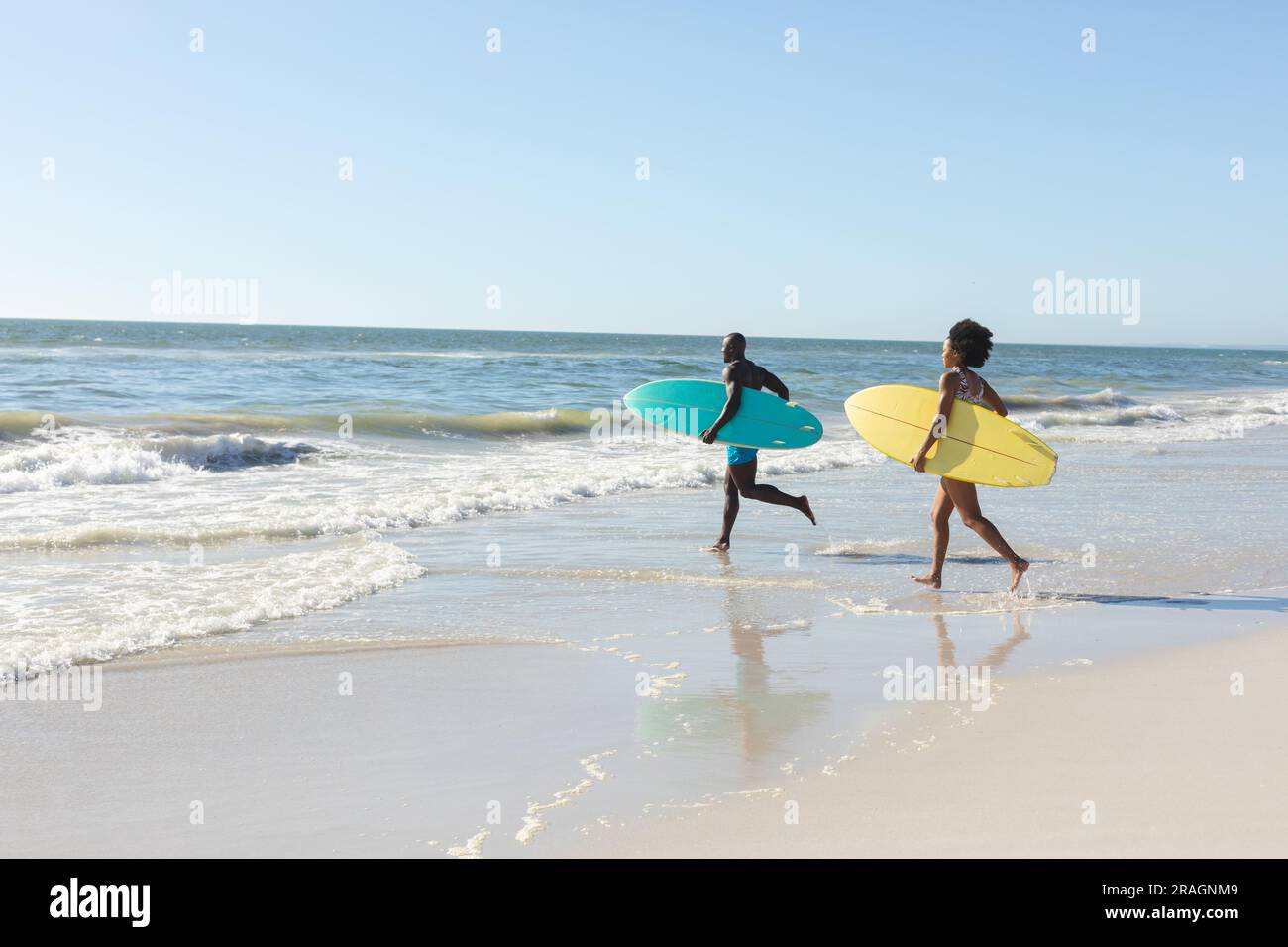 African american couple carrying surfboards running on sunny beach to ...