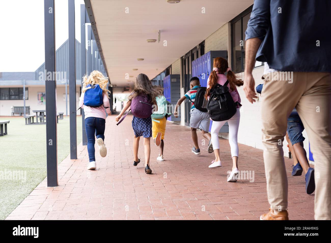 Rear view of diverse male teacher with children running in elementary ...