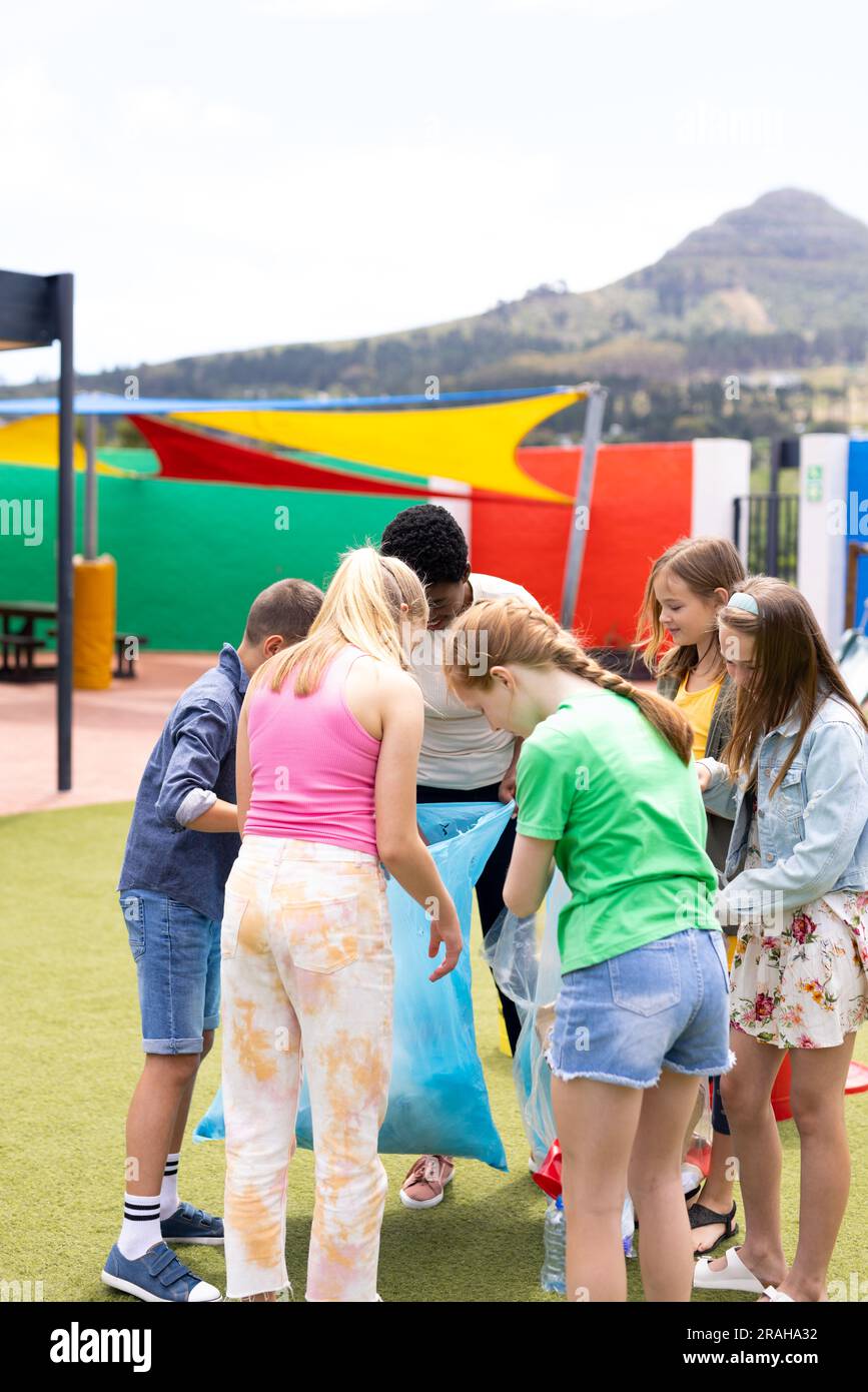 Vertical of diverse female teacher and pupils collecting recycling in ...
