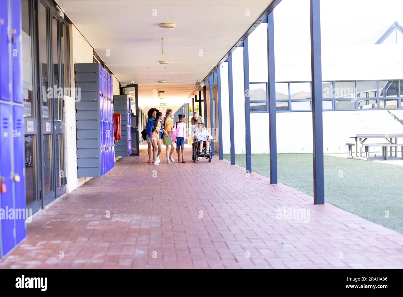 Diverse group of school children walking in school corridor talking, one in wheelchair, copy ...