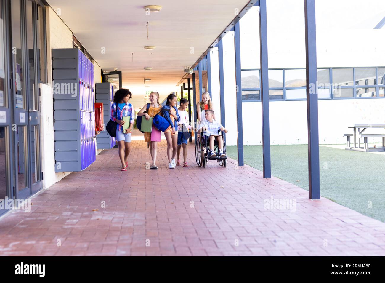 Diverse group of school children walking in school corridor talking, one in wheelchair, copy ...