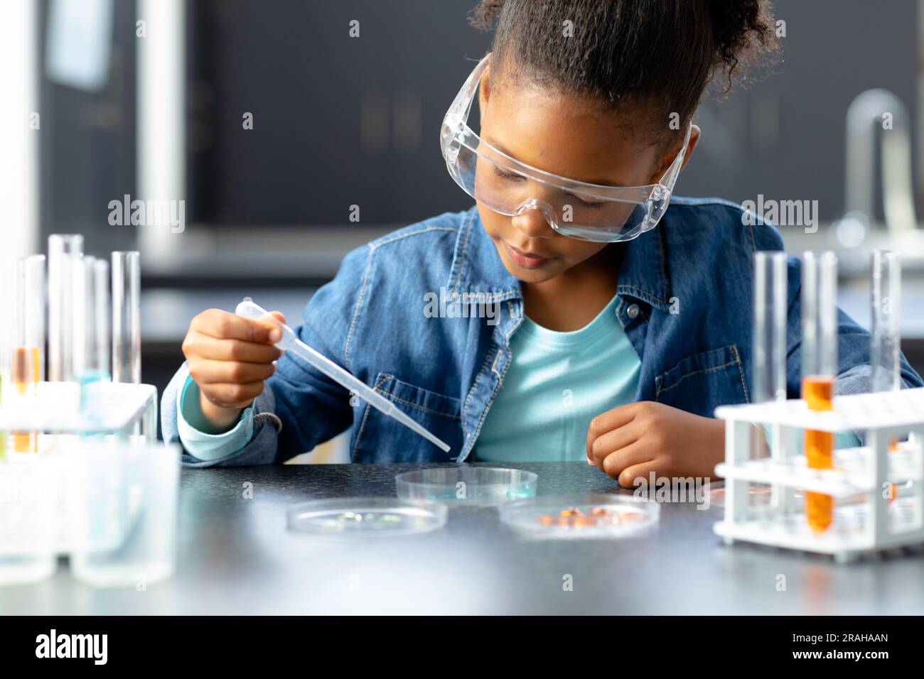 Happy biracial schoolgirl in safety glasses using pipette in science ...